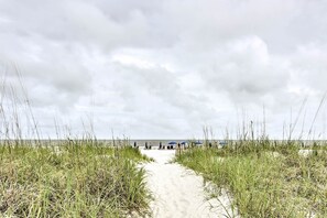 Um caminho de areia ao longo da praia, ladeado por grama alta, leva até um cais com guarda-sóis.