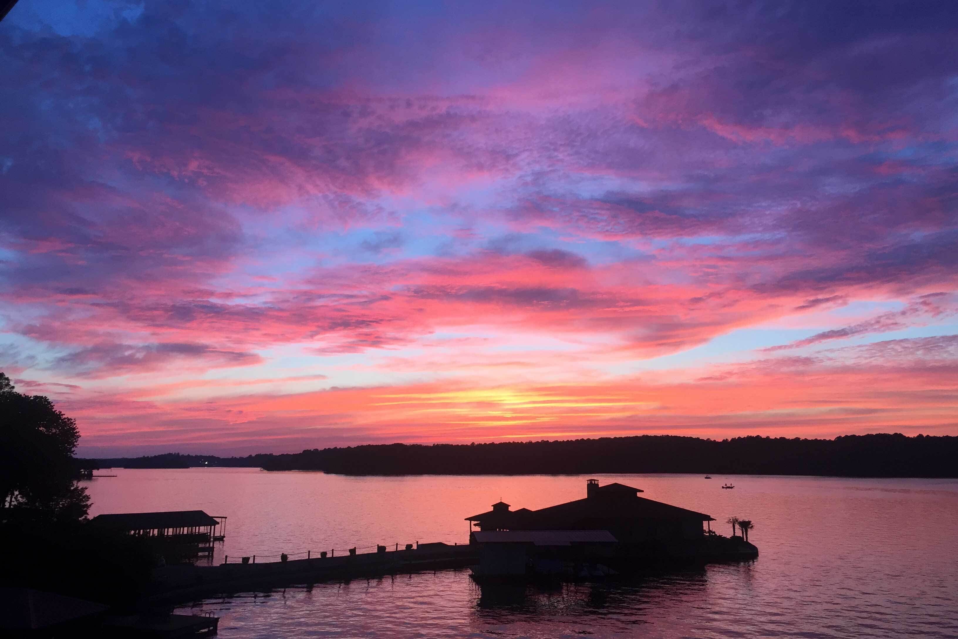 Um pôr do sol sobre um lago calmo, com um píer e edifícios na margem.