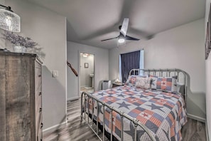 Quarto com estrutura de cama de metal, colcha acolchoada, ventoinha no teto e cómoda de madeira.
