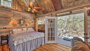 Quarto rústico com teto de madeira, cama com roupa de cama florida, ventilador de teto e vista para o exterior.