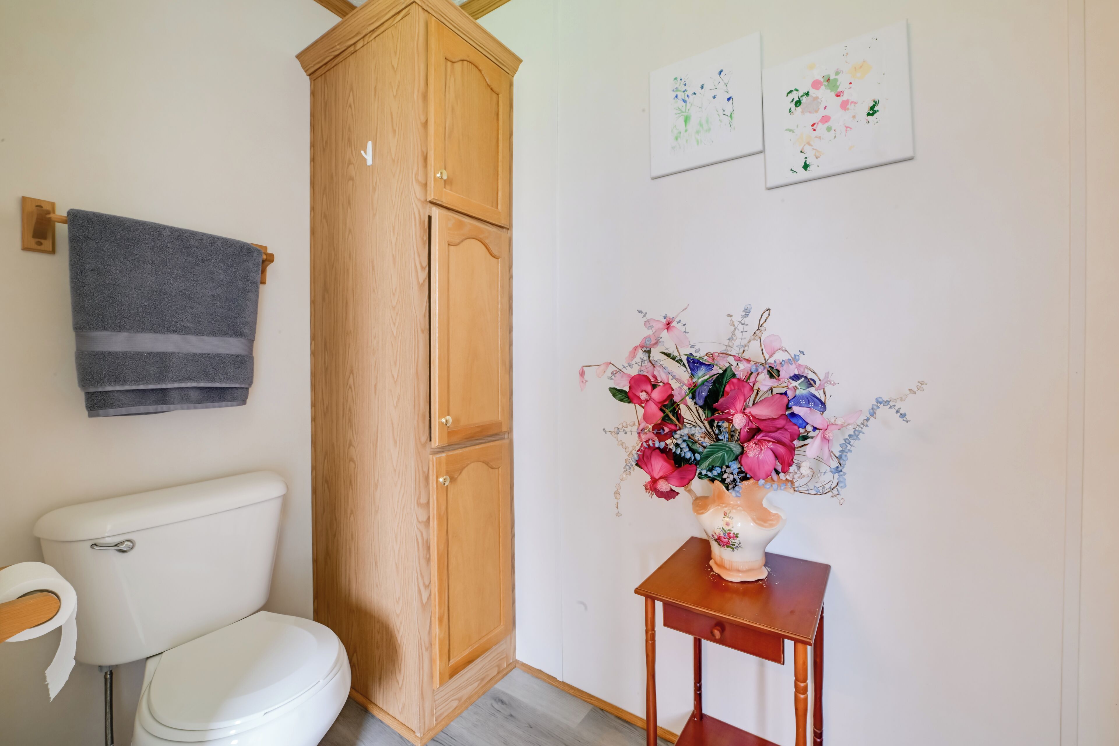 Casa de banho com sanita, armário de madeira e um vaso de flores sobre uma pequena mesa.