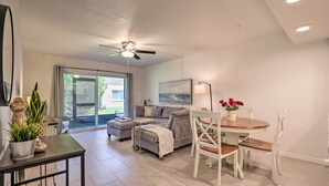 Uma sala de estar moderna com sofá, pufê, mesa de jantar e cadeiras. Há um ventilador de teto, uma janela e um quadro na parede.
