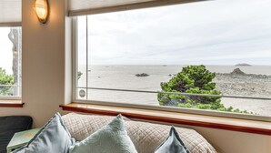 Quarto com uma grande janela com vista para uma praia, uma cama com almofadas e um candeeiro fixo na parede.
