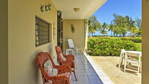 Área de estar coberta ao ar livre, com cadeiras de vime e uma mesa, com vista para um jardim paisagístico e palmeiras.