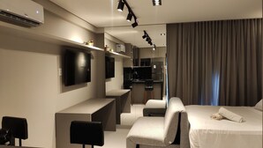 Quarto de hotel moderno com televisão de tela plana, pequena cozinha, área de estar com um banco e uma janela grande com cortinas.