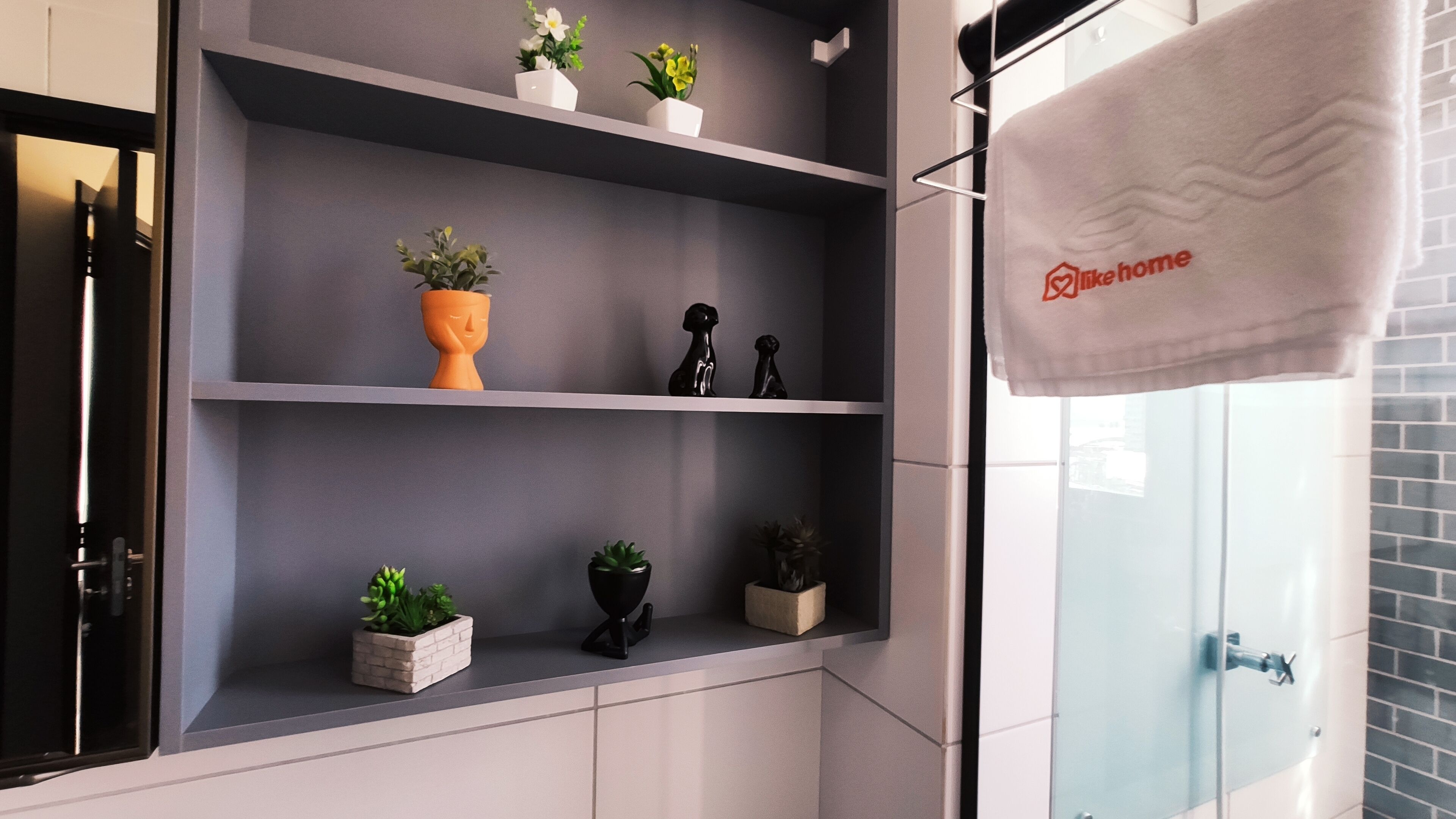 Casa de banho moderna com prateleiras cinzentas com objetos decorativos, um porta-toalhas com toalha que transmite a sensação de lar, e uma porta de cabine de duche em vidro.