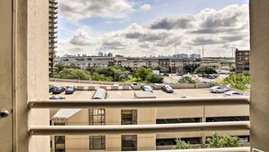 Vista de uma varanda sobre um parque de estacionamento com vários carros, paisagem urbana ao fundo com edifícios e céu parcialmente nublado.