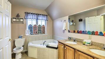Um banheiro com vaso sanitário, banheira, duas pias, armários de madeira, um espelho grande e uma janela com vista para casas coloridas.