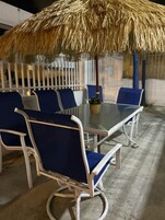 Uma área de refeições com mesa de vidro e cadeiras azuis, um guarda-sol de palha e uma cerca branca ao fundo.