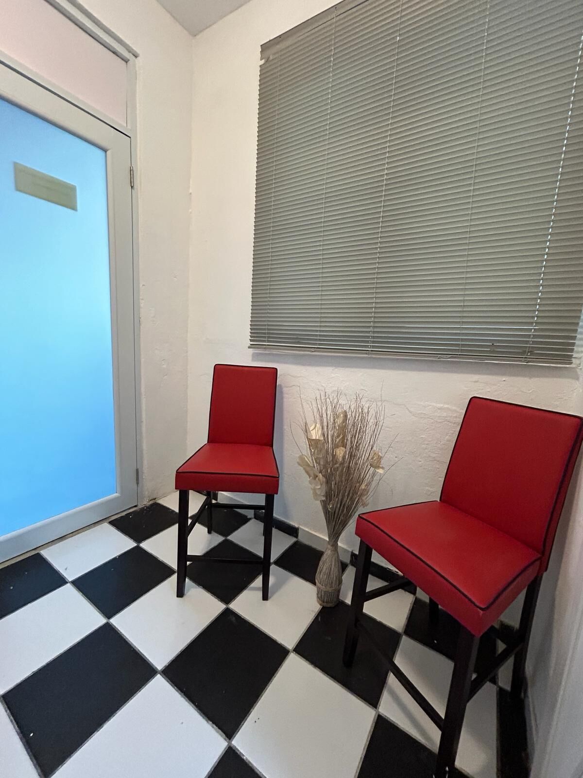 Um quarto com chão quadriculado, duas cadeiras vermelhas, um vaso com flores secas, uma janela com persianas e uma porta.