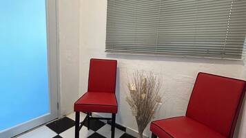 Um quarto com chão quadriculado, duas cadeiras vermelhas, um vaso com flores secas, uma janela com persianas e uma porta.