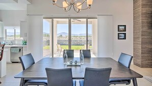 Uma área de jantar moderna com uma mesa grande, cadeiras e um candelabro. Vê-se um pátio com mesas e cadeiras ao ar livre e uma paisagem montanhosa ao fundo.