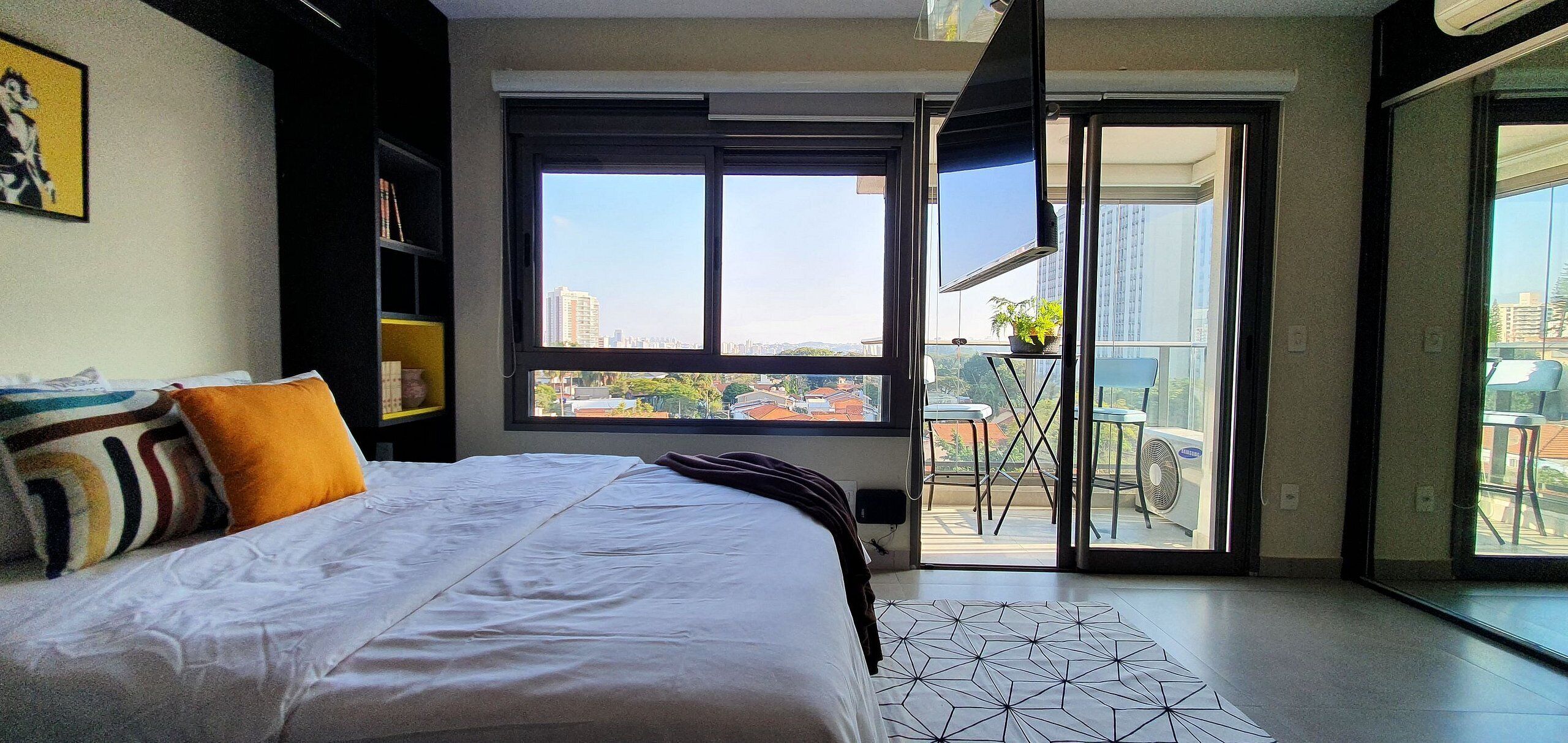 Quarto moderno com cama grande, vista da cidade através de uma porta de vidro deslizante e varanda com mobiliário exterior.