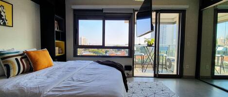 Quarto moderno com cama grande, vista da cidade através de uma porta de vidro deslizante e varanda com mobiliário exterior.