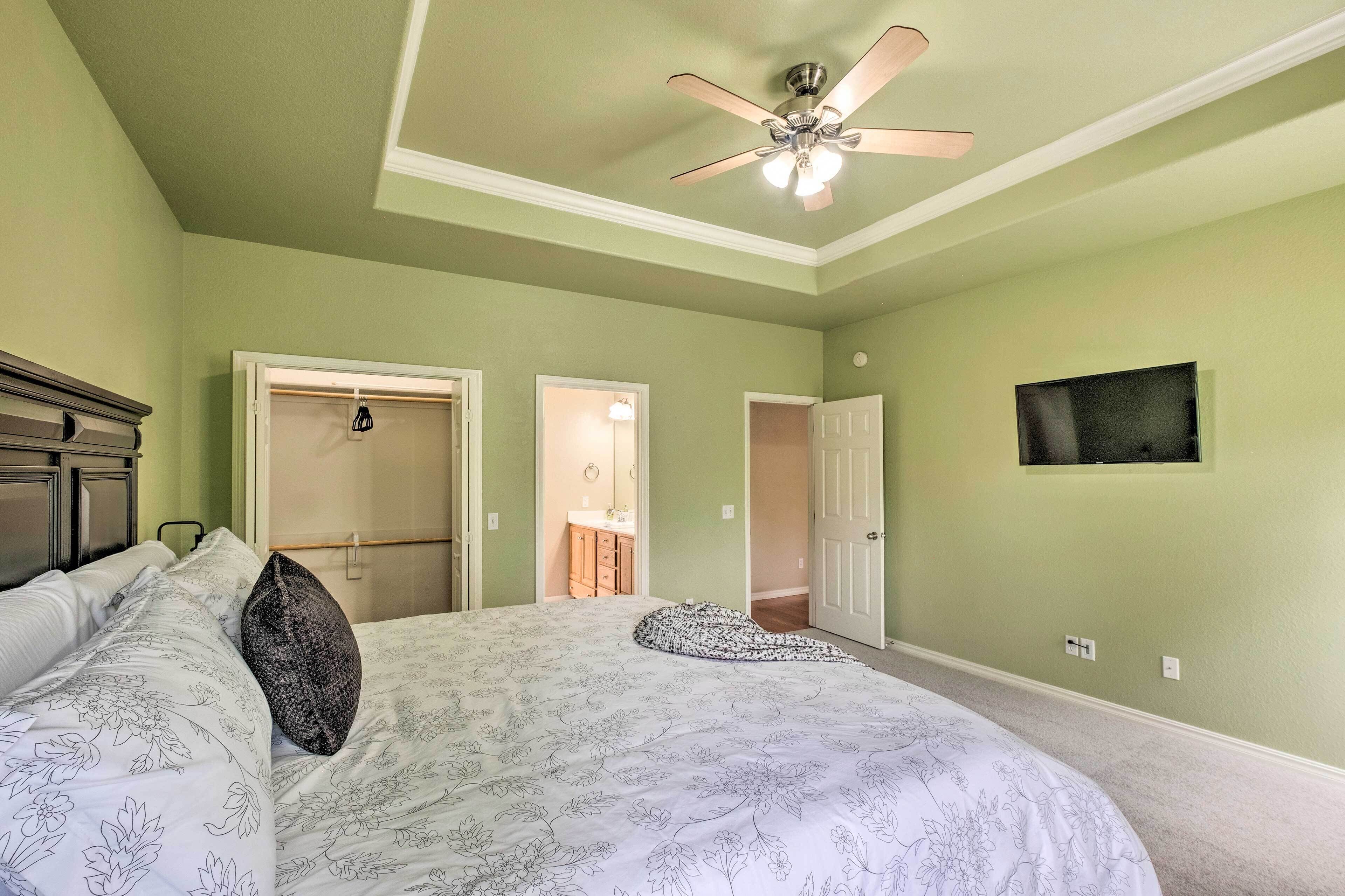 Quarto com cama, ventilador de teto, TV fixada na parede e porta que dá para outro cômodo.