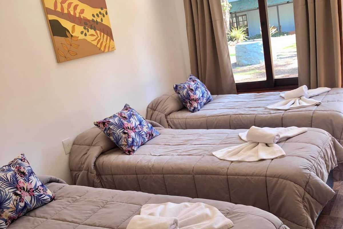 Quarto de hotel com três camas, cada uma com um edredom bege e almofada florida. Há um quadro na parede e uma janela com cortinas.