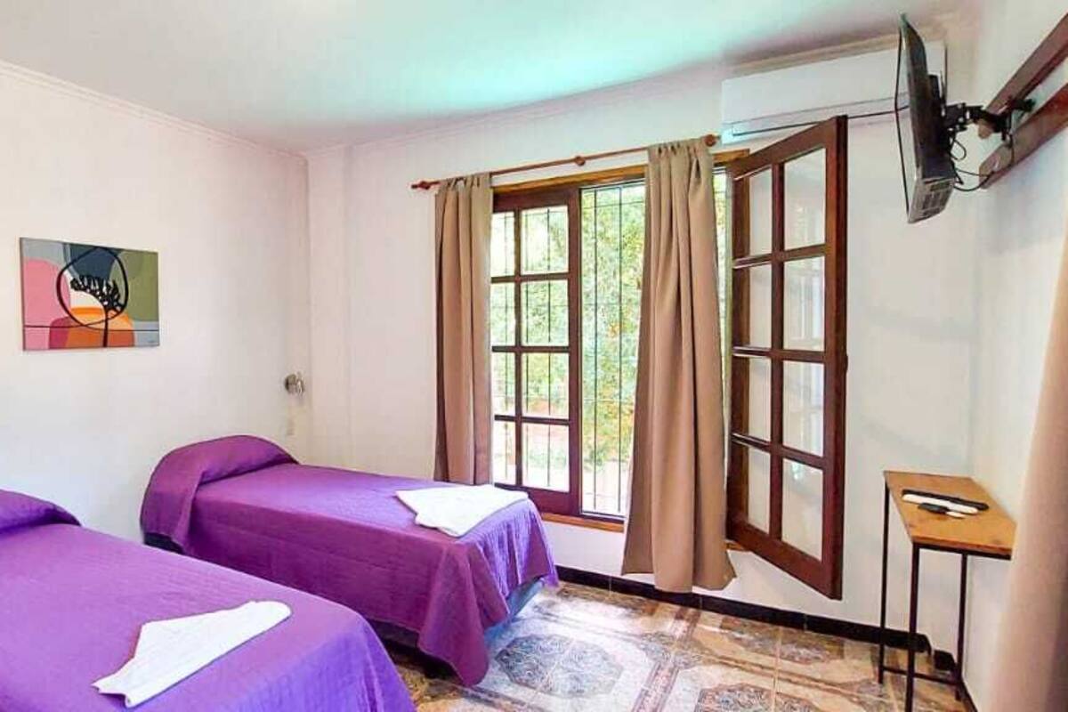 Quarto com duas camas, janela com cortinas, televisão na parede e uma pequena mesa.