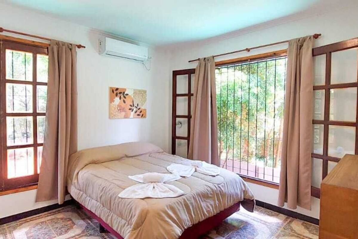 Quarto com cama, janelas amplas, cabeceira de madeira e ar condicionado fixado na parede.