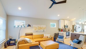 Uma sala de estar moderna com sofá, um banquinho e poltronas, uma cozinha equipada e um ventilador de teto.