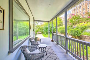 Varanda coberta com cadeiras de vime e uma pequena mesa, com vista para um jardim e um edifício de tijolos.