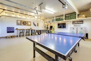 Mesa de pingue-pongue em um quarto com alvo de dardos, uma placa com a inscrição 'GAME ROOM' e prateleiras com cobertores dobrados.