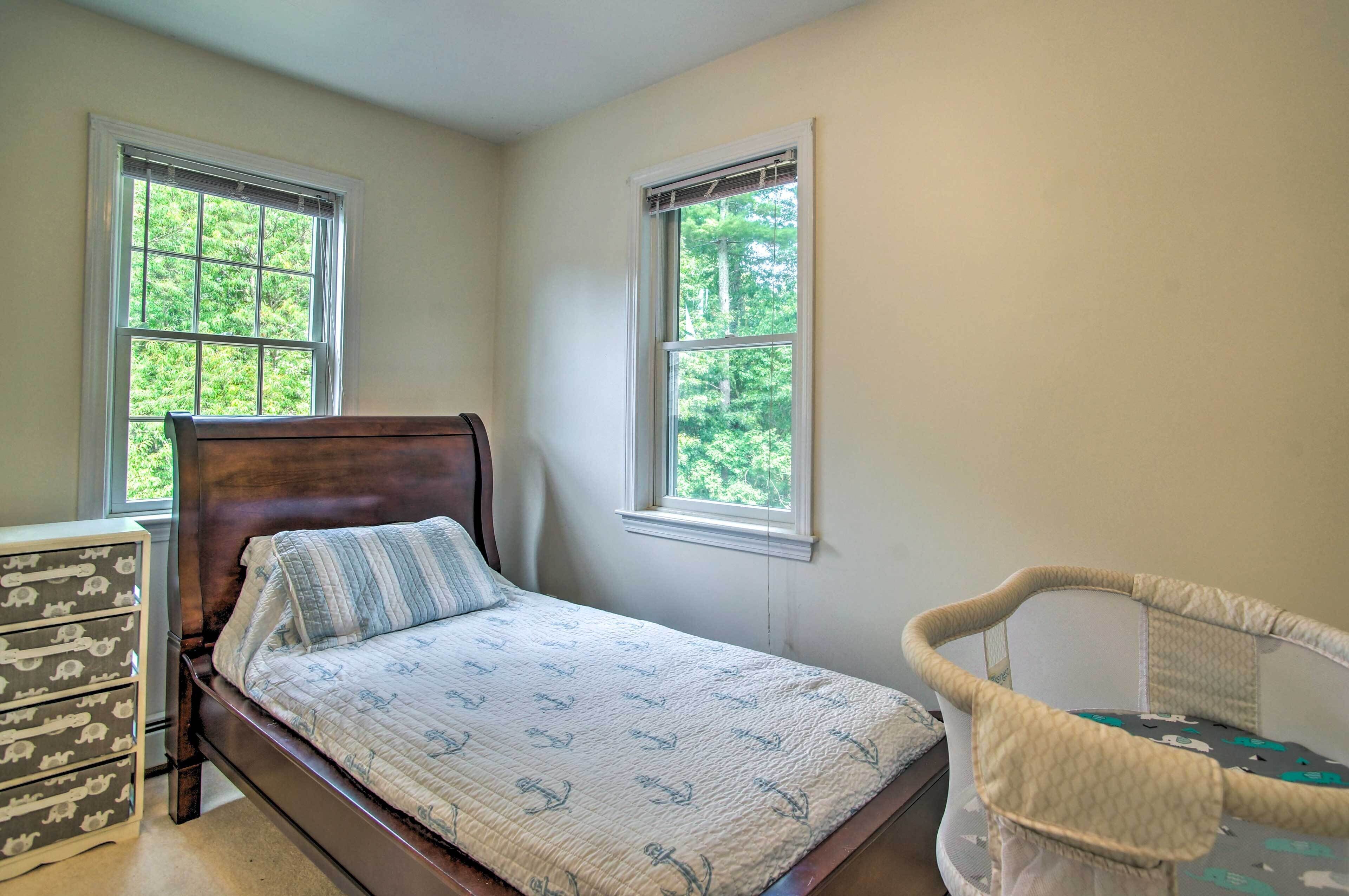 Quarto com cama de madeira, duas janelas, berço e cómoda com gavetas.