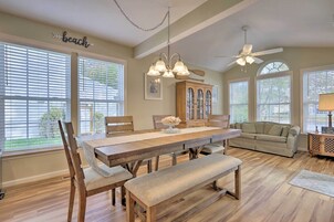 Uma sala de jantar com uma mesa de madeira, bancos e um lustre. Há janelas amplas com persianas, um ventilador de teto e um armário de madeira.