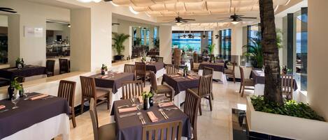 Um restaurante com várias mesas, ventiladores de teto e grandes janelas com vista para o oceano.