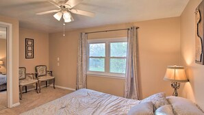 Quarto com cama, ventoinha no teto, janela com cortinas, duas cadeiras e mesa de cabeceira com abajur.