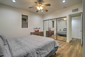 Um quarto com cama, ventilador de teto, cômoda, espelho e uma porta deslizante que dá para outro cômodo.