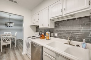 Cozinha com armários brancos, lava-louças de aço inoxidável, pia branca e painel de azulejos cinza.
