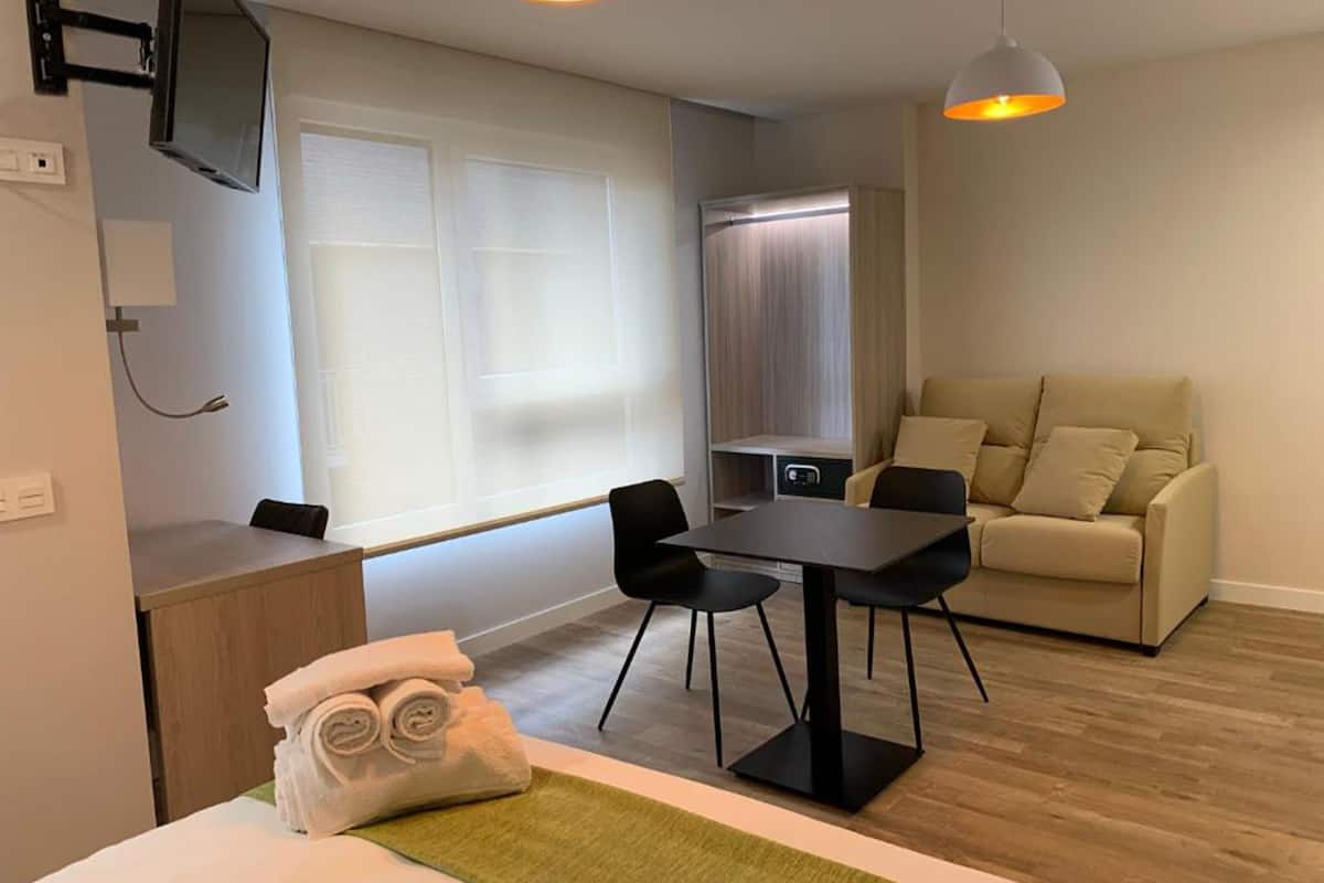 Quarto de hotel moderno com uma cama, uma escrivaninha, uma mesa de jantar com cadeiras, um sofá e uma TV fixada na parede.