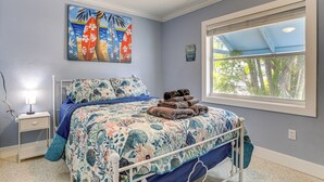 Quarto com cama, mesa de cabeceira, ventilador de teto, janela com vista para árvores e pintura de pranchas de surf.