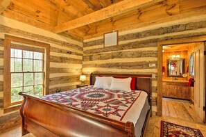 Quarto de cabana de madeira com cama de madeira, janela com persianas, mesa de cabeceira com abajur e banheiro visível por uma porta aberta.