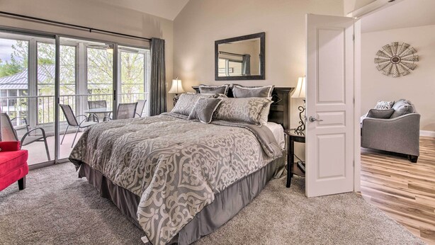 Quarto com uma cama grande, ventilador de teto, janela com cortinas, mesa de cabeceira com abajur e porta para outro cômodo.