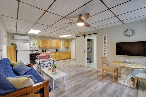 Uma sala de estar ampla com sofá, mesa de centro e uma área de refeições com mesa e cadeiras. Dá para ver uma cozinha ao fundo, com armários de madeira e uma geladeira.
