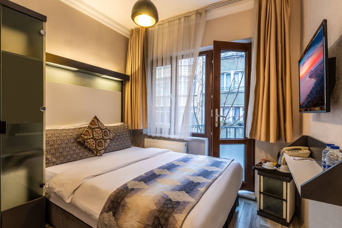 Quarto de hotel com uma cama, uma escrivaninha com telefone e uma garrafa, uma televisão na parede e uma janela com cortinas.