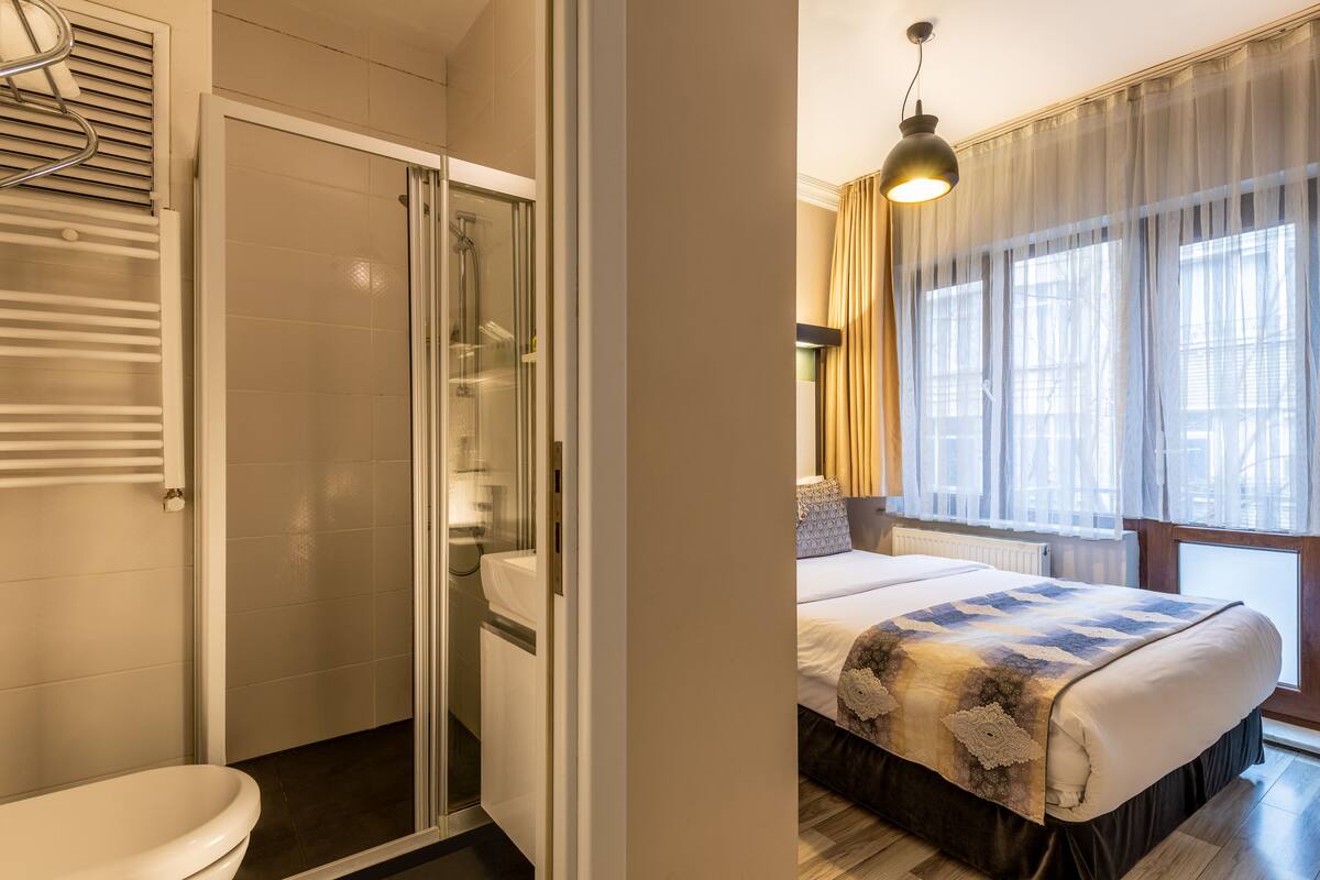 Quarto de hotel com cabine de chuveiro de vidro, cama com manta estampada e grande janela com cortinas.