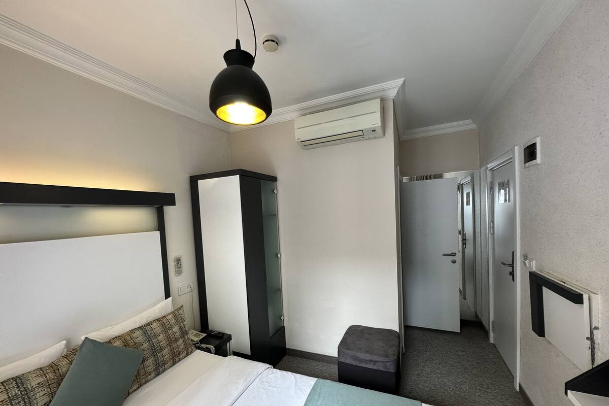 Quarto de hotel com cama, mesa de cabeceira, abajur, guarda-roupa e porta para um corredor.