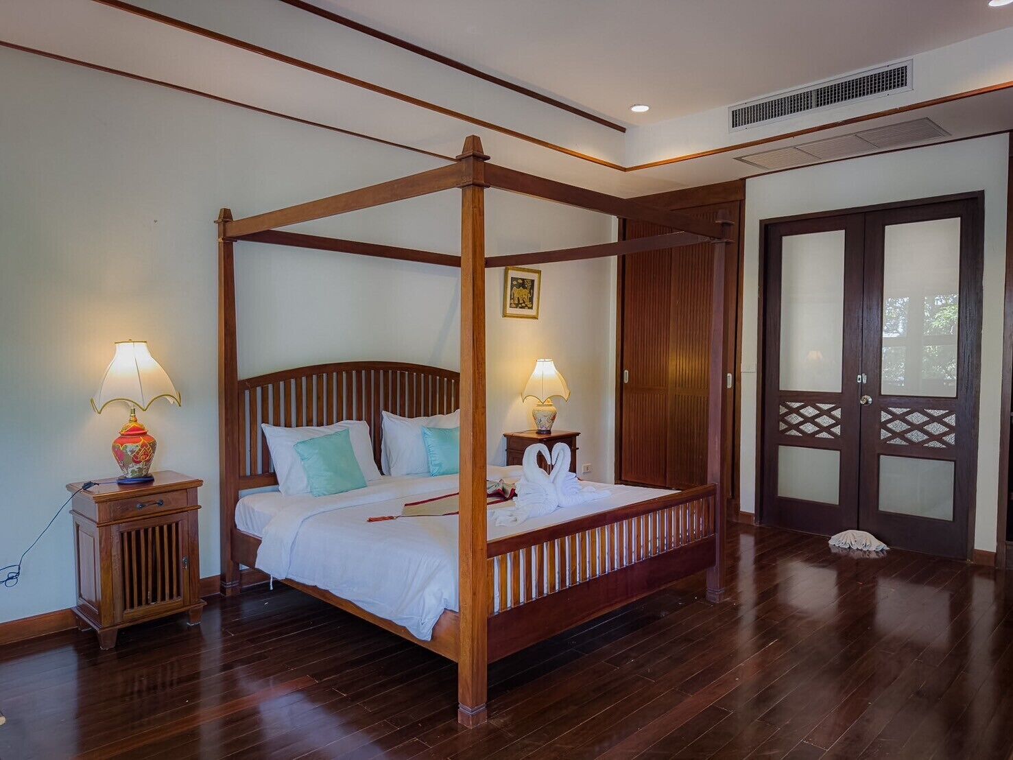 Uma cama de dossel com estrutura de madeira e roupa de cama branca, duas luminárias de cabeceira, um pequeno armário e uma porta de madeira com painéis de vidro.
