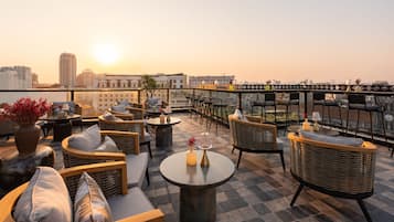 Terraço no telhado com móveis de vime, mesas redondas e vista para o horizonte urbano ao pôr do sol.