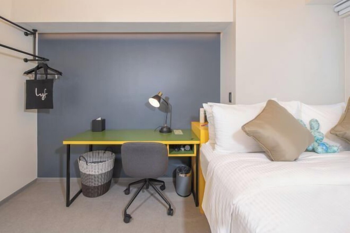 Quarto de hotel pequeno com cama, escrivaninha, cadeira, abajur, lixeira e luminária na parede.