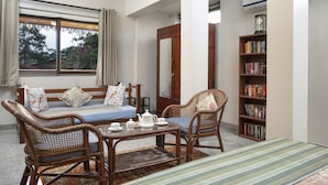 Quarto com cama, duas cadeiras de vime, uma mesinha com um bule, uma estante de livros e uma janela com cortinas.