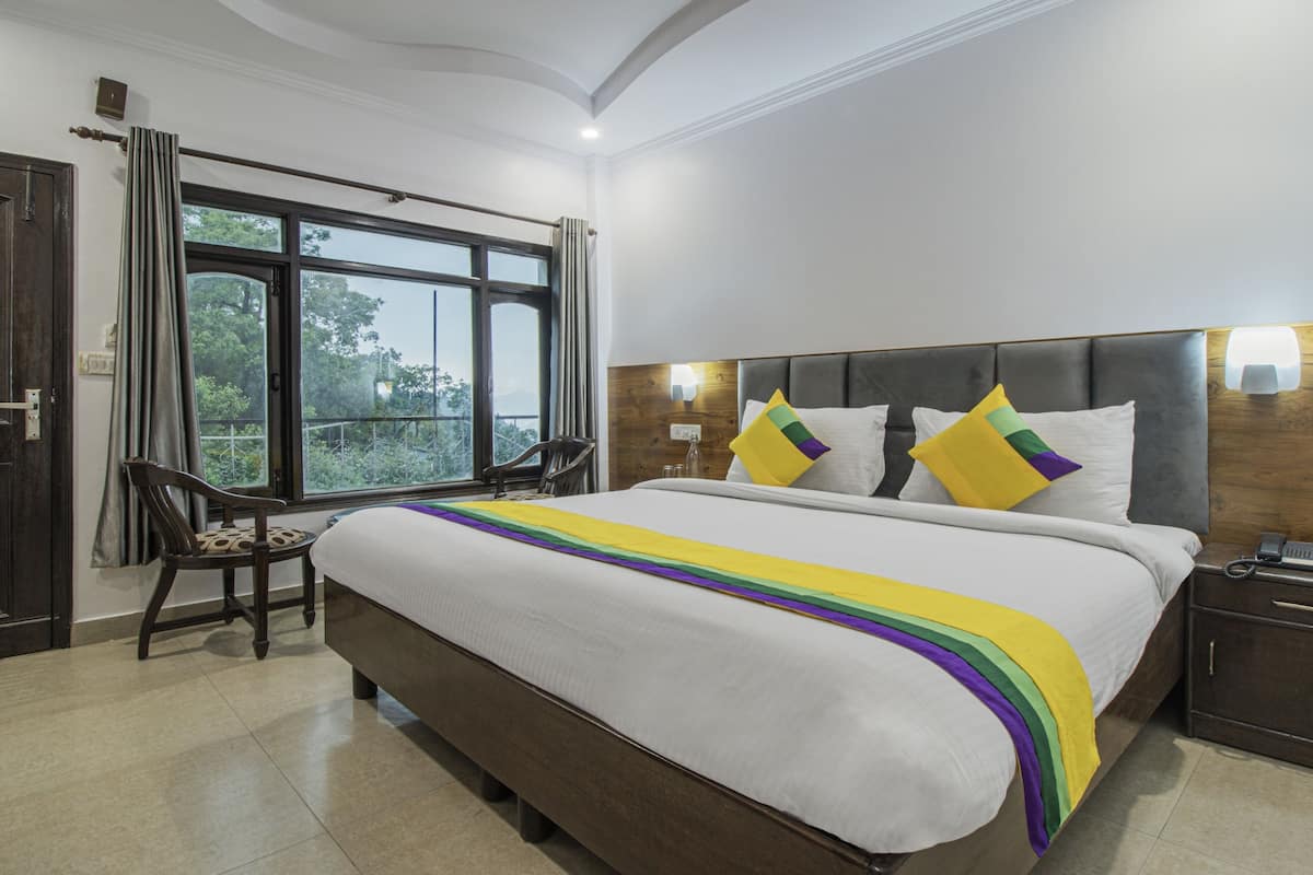 Quarto de hotel com uma cama grande, cabeceira de madeira, mesa de cabeceira com telefone e janela com cortinas.