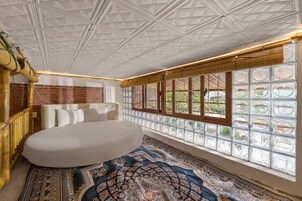 Quarto com cama grande, tapete estampado, parede de tijolos e janela com vista para área verde.