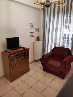 Uma sala de estar com um armário de madeira, uma televisão, um sofá estampado, um radiador e uma janela com cortinas.