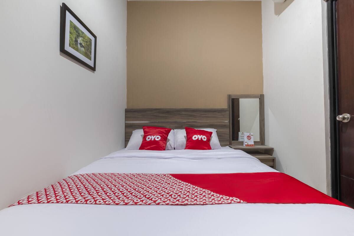Quarto de hotel com uma cama, duas almofadas com a marca 'OYO', um edredom com padrão vermelho e branco, um quadro na parede e uma cabeceira de madeira.