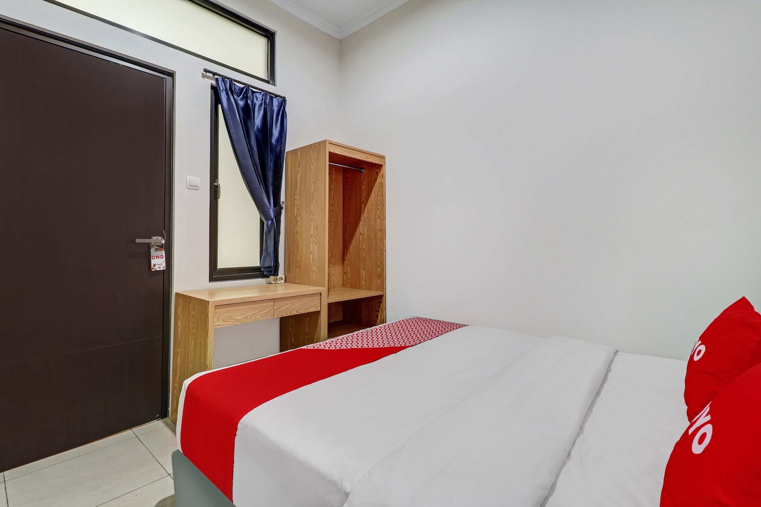 Quarto de hotel com uma cama, uma escrivaninha de madeira, um guarda-roupa e uma porta.