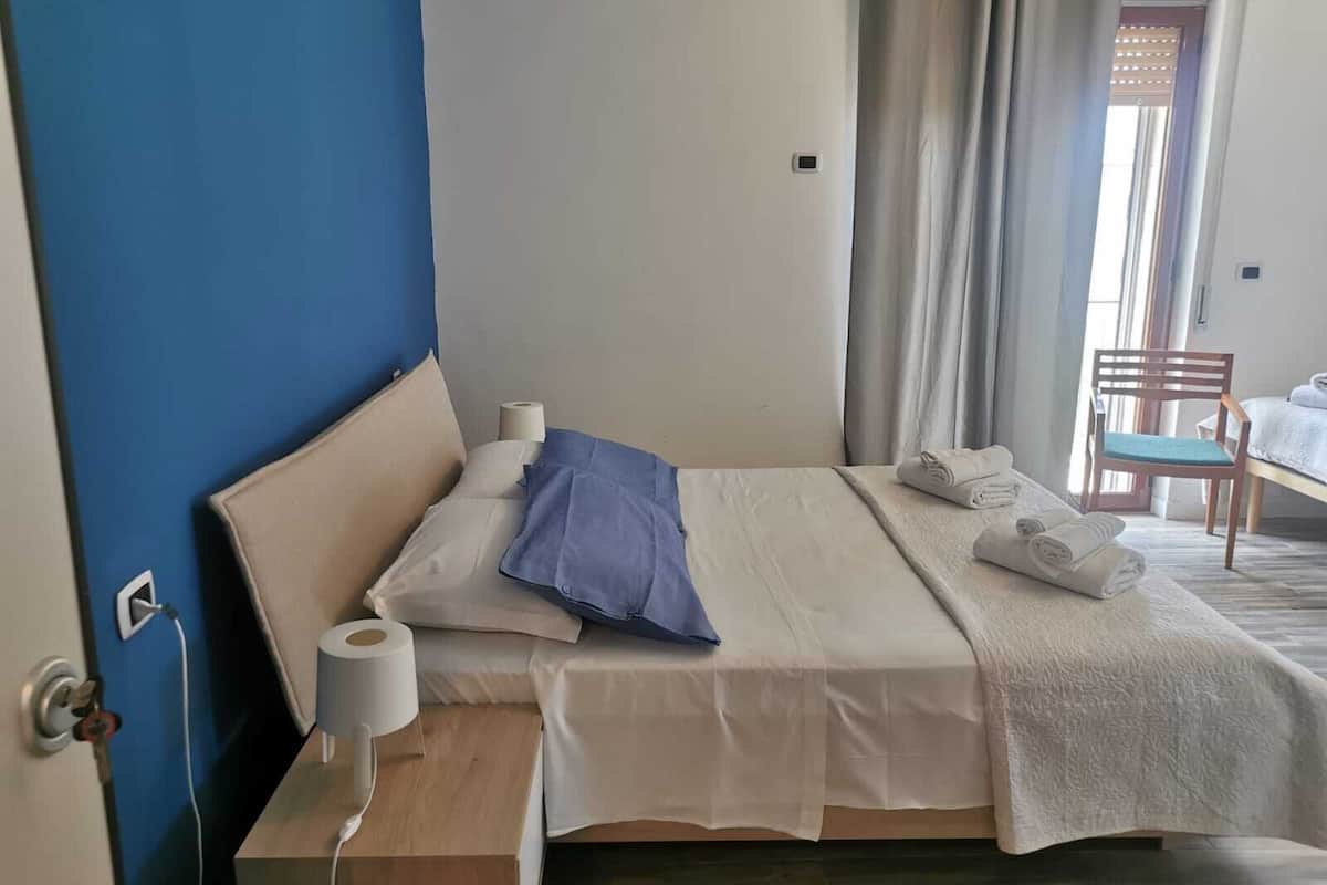 Quarto de hotel com cama, mesa de cabeceira de madeira, uma cadeira e janela com cortinas.