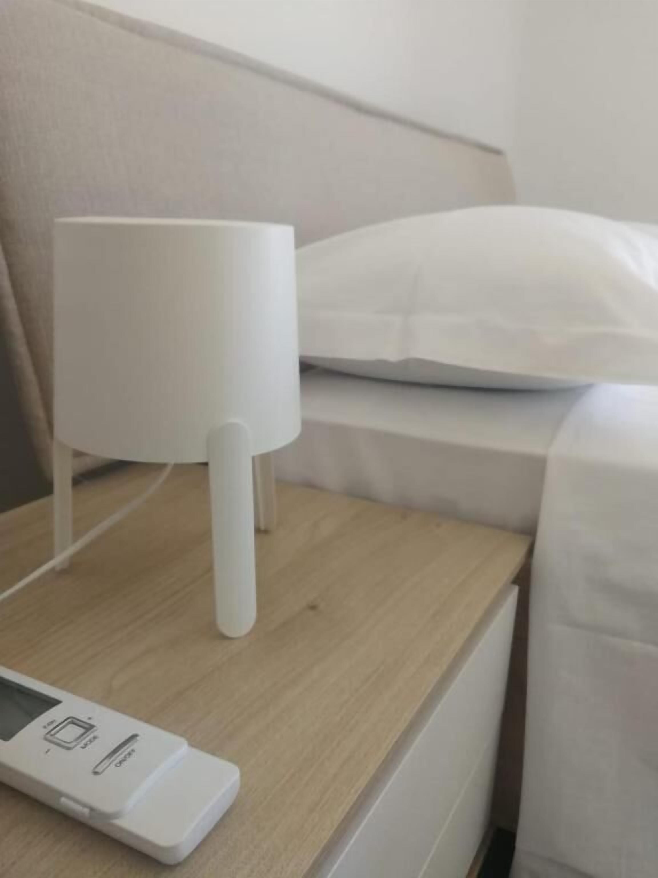 Uma mesa de cabeceira branca com uma lâmpada e um dispositivo digital, provavelmente um termostato, em um quarto de hotel.